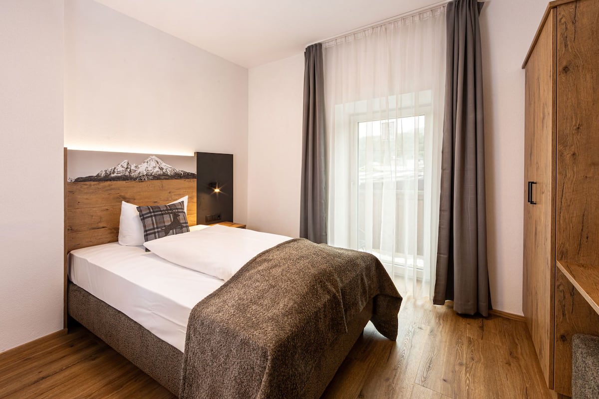 Quarto com cabeceira de madeira, cama com roupa de cama branca, mesa de cabeceira, luminária fixada na parede e vista para uma montanha através de uma janela com cortinas.
