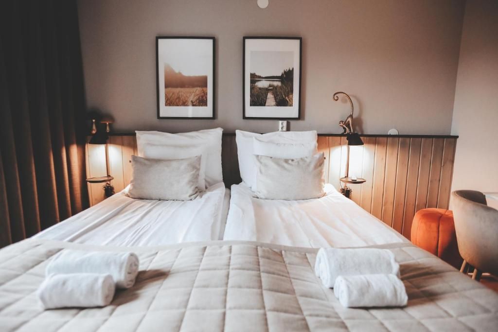 Uma cama de casal com lençóis brancos e duas almofadas, uma mesa de cabeceira com abajur e duas quadros decorativos na parede.