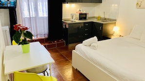 Quarto de hotel pequeno com uma cama, uma kitchenette, uma mesa de jantar com uma cadeira e uma TV fixada na parede.