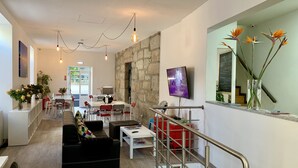 Uma sala de estar moderna com um sofá preto, mesa de jantar branca e cadeiras vermelhas. Há uma televisão fixada na parede, uma parede de pedra e uma escada com corrimão de metal.
