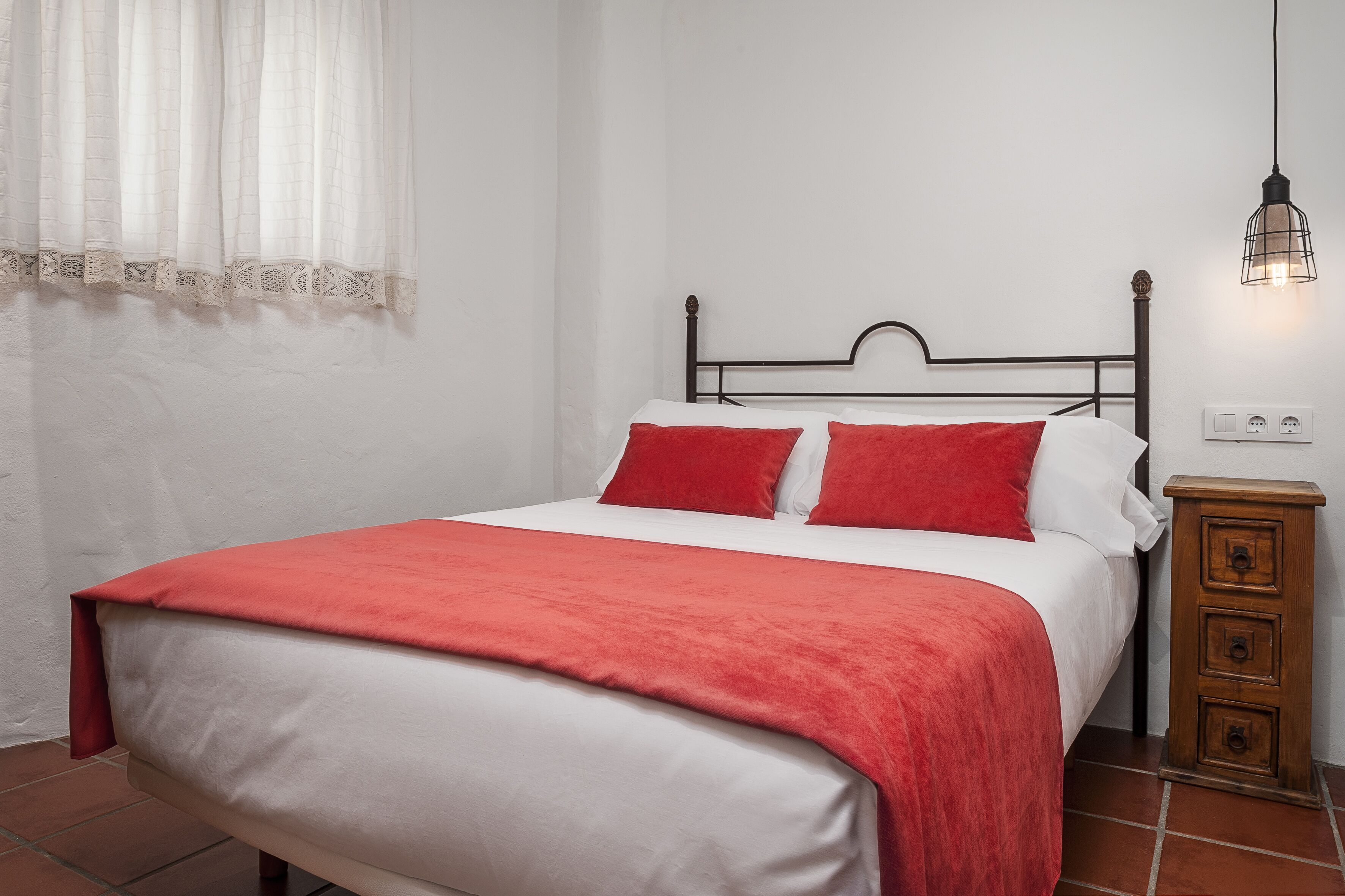 Uma cama com edredom branco e vermelho, uma mesa de cabeceira de madeira com gavetas e um aparelho de luz fixado na parede.