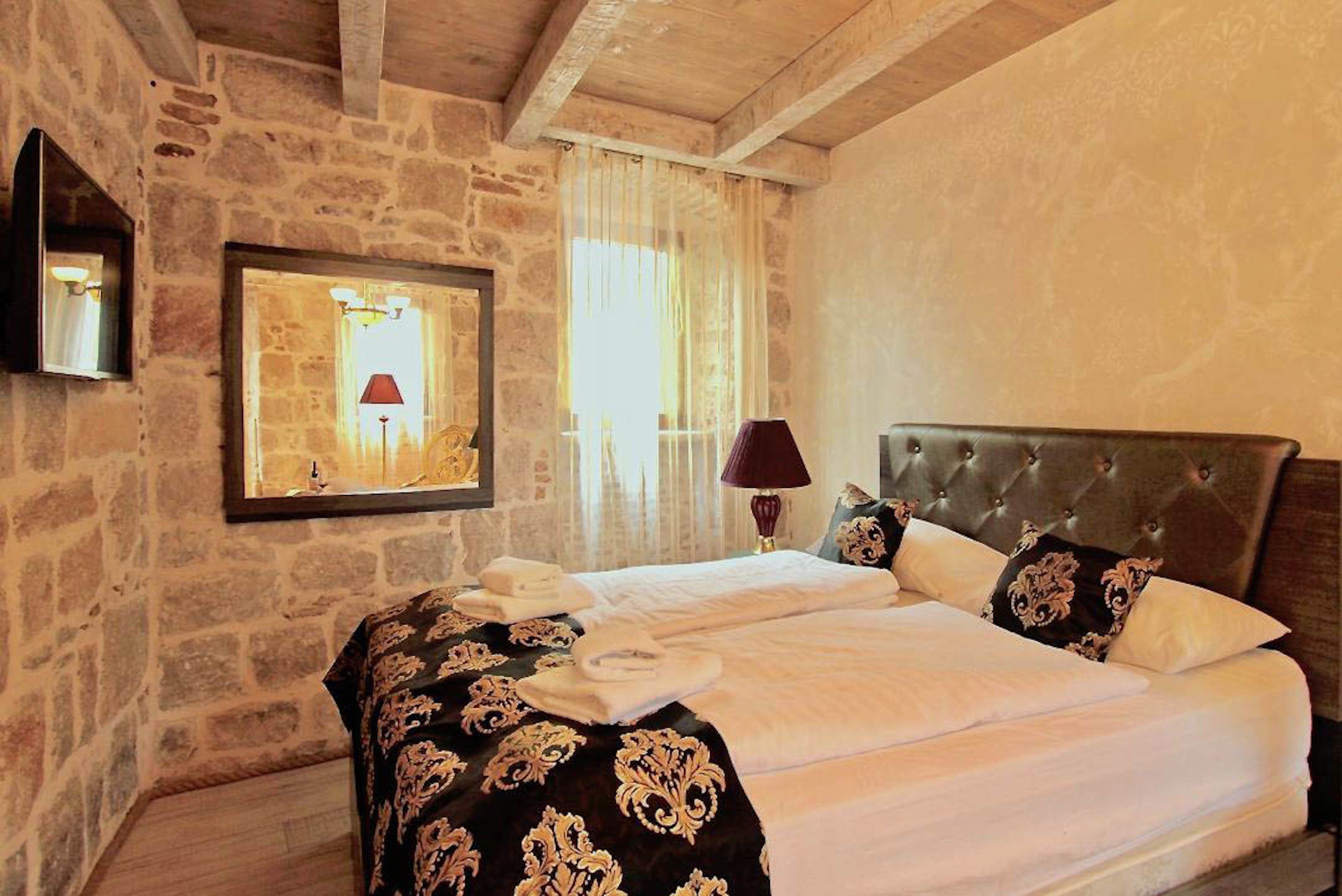 Quarto com parede de pedra, cama com colcha estampada escura, cabeceira estofada e quadro pendurado na parede.