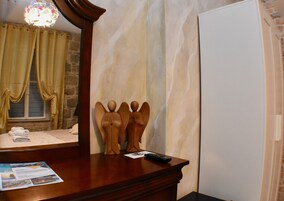Quarto com cómoda de madeira, espelho, escultura de anjo de madeira, comando de televisor e janela com cortinas.