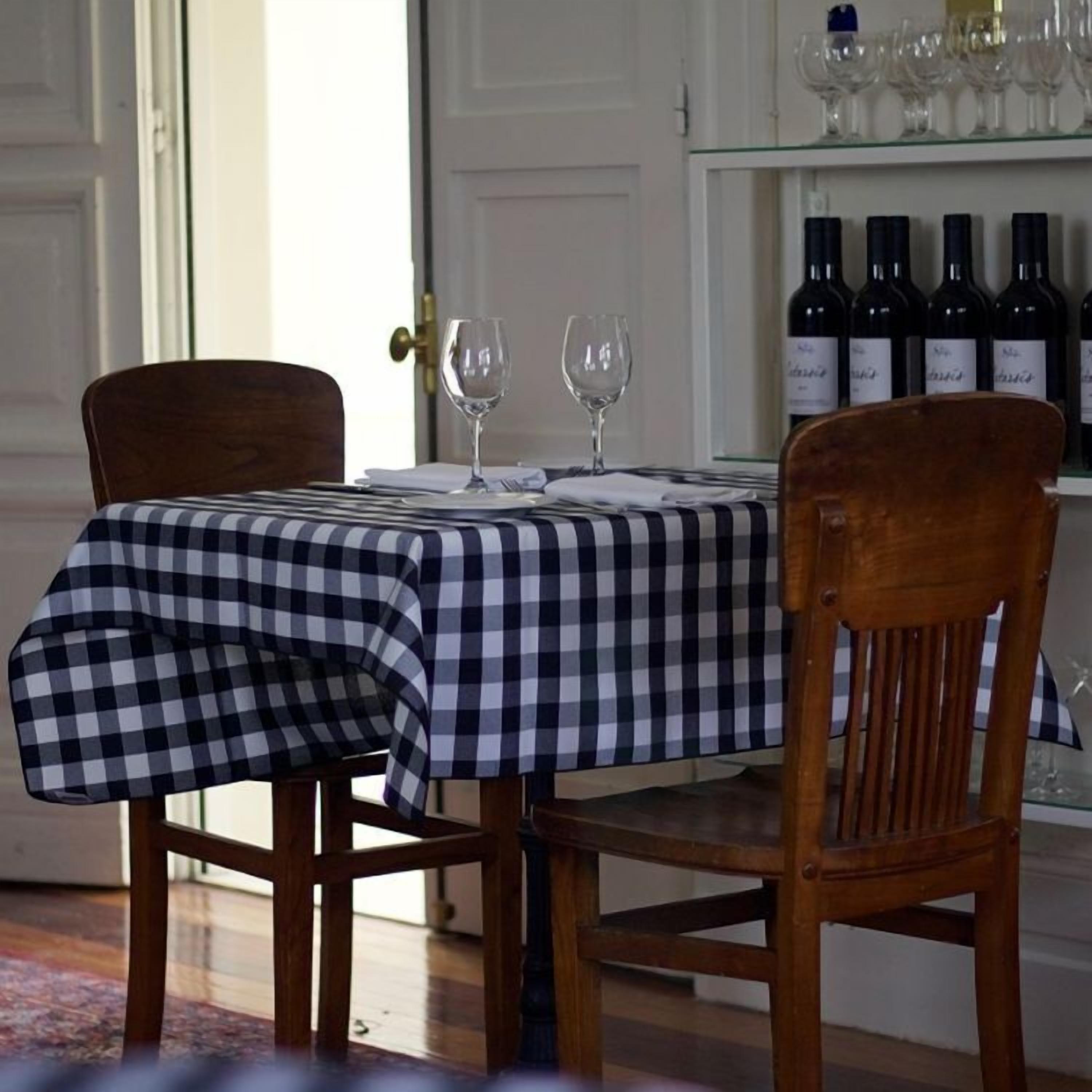 Mesa de jantar com toalha xadrez, duas taças de vinho e uma garrafa de vinho sobre ela. Duas cadeiras de madeira estão dispostas ao redor da mesa. Ao fundo, há uma estante com diversas garrafas de vinho.