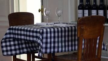 Mesa de jantar com toalha xadrez, duas taças de vinho e uma garrafa de vinho sobre ela. Duas cadeiras de madeira estão dispostas ao redor da mesa. Ao fundo, há uma estante com diversas garrafas de vinho.