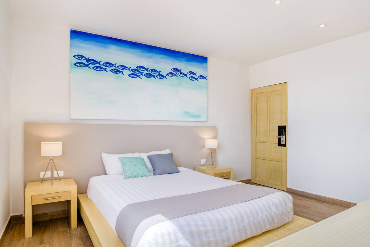 Quarto moderno com uma cama grande, duas mesas de cabeceira com abajures, um guarda-roupa de madeira e uma pintura de peixes na parede.