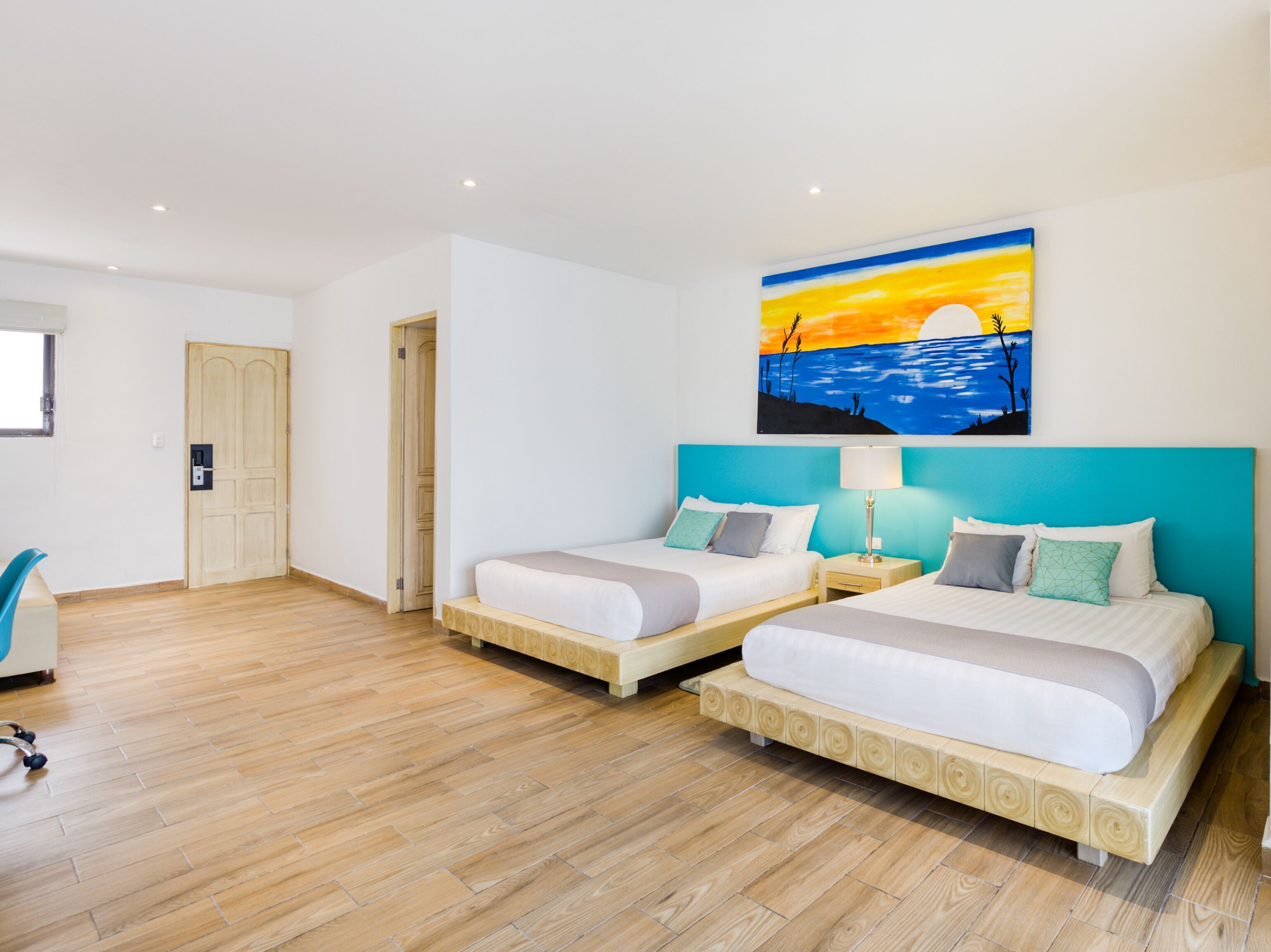 Quarto de hotel com duas camas, uma pintura de um pôr do sol e piso de madeira.