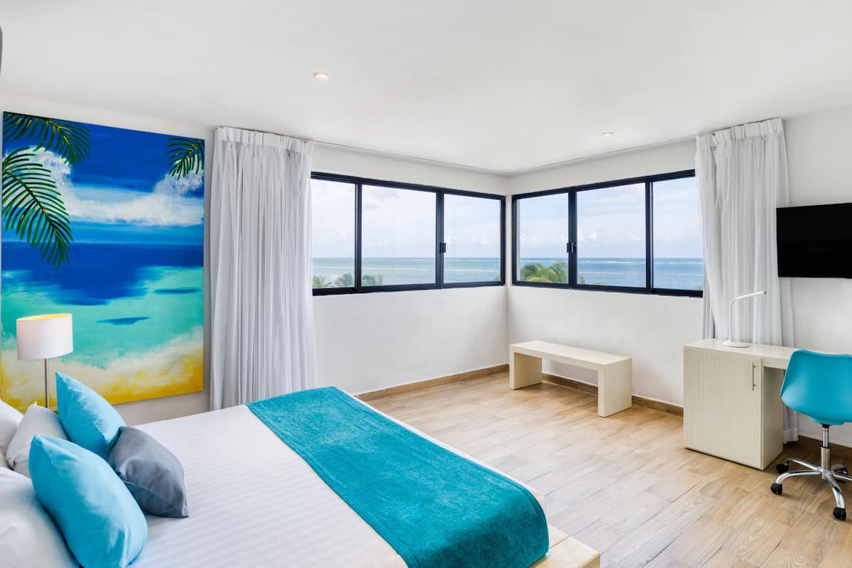 Quarto de hotel moderno com uma cama grande, uma mesa de trabalho, uma cadeira e uma televisão. O quarto possui amplas janelas com vista para o oceano.