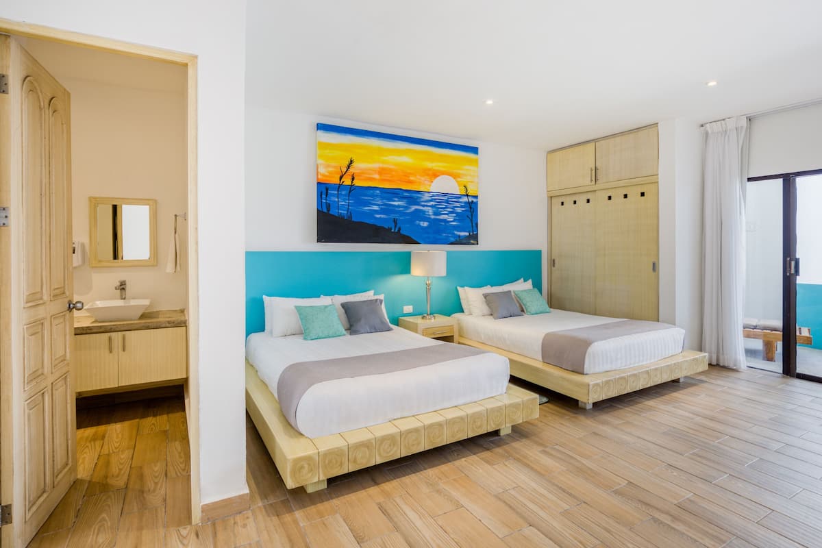 Quarto de hotel com duas camas, um banheiro, uma pintura do pôr do sol e vista para o mar.