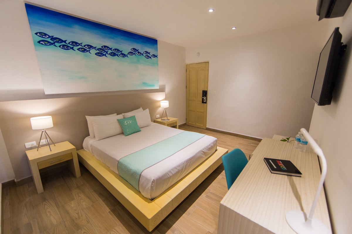 Quarto de hotel com cama, mesas de cabeceira, escrivaninha, cadeira, quadro e porta.