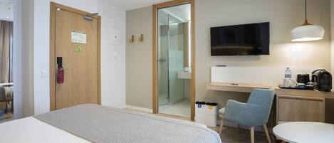 Quarto de hotel com cama, escrivaninha, cadeira, televisão e zona de chuveiro.