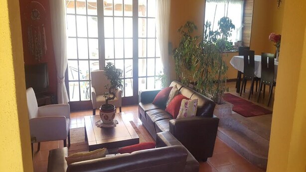 Uma sala de estar com sofá, poltronas, mesa de centro e televisão. Ao fundo, uma área de jantar com mesa e cadeiras.