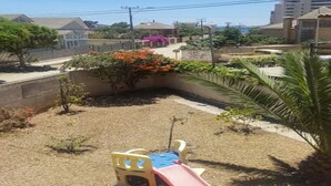 Uma área residencial com um caminho de entrada pavimentado, um pequeno jardim com uma palmeira e uma cadeira amarela.