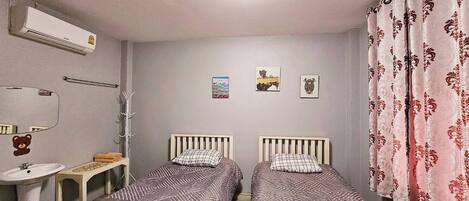 Quarto pequeno e bem iluminado, com duas camas de solteiro, uma mesa de cabeceira, um espelho, ar-condicionado fixado na parede e uma janela com cortinas.