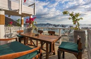 Mesa e cadeiras de madeira com almofadas verdes, cerca branca e vista para barcos na água.