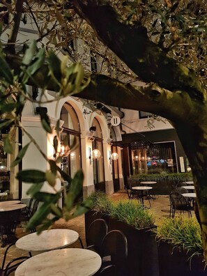 Uma área de café ao ar livre, com mesas e cadeiras, cercada por vegetação e arcos arquitetônicos.