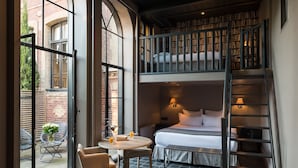 Quarto de hotel com uma cama grande, varanda com cadeiras, uma mesinha com bebidas e uma estante de livros.