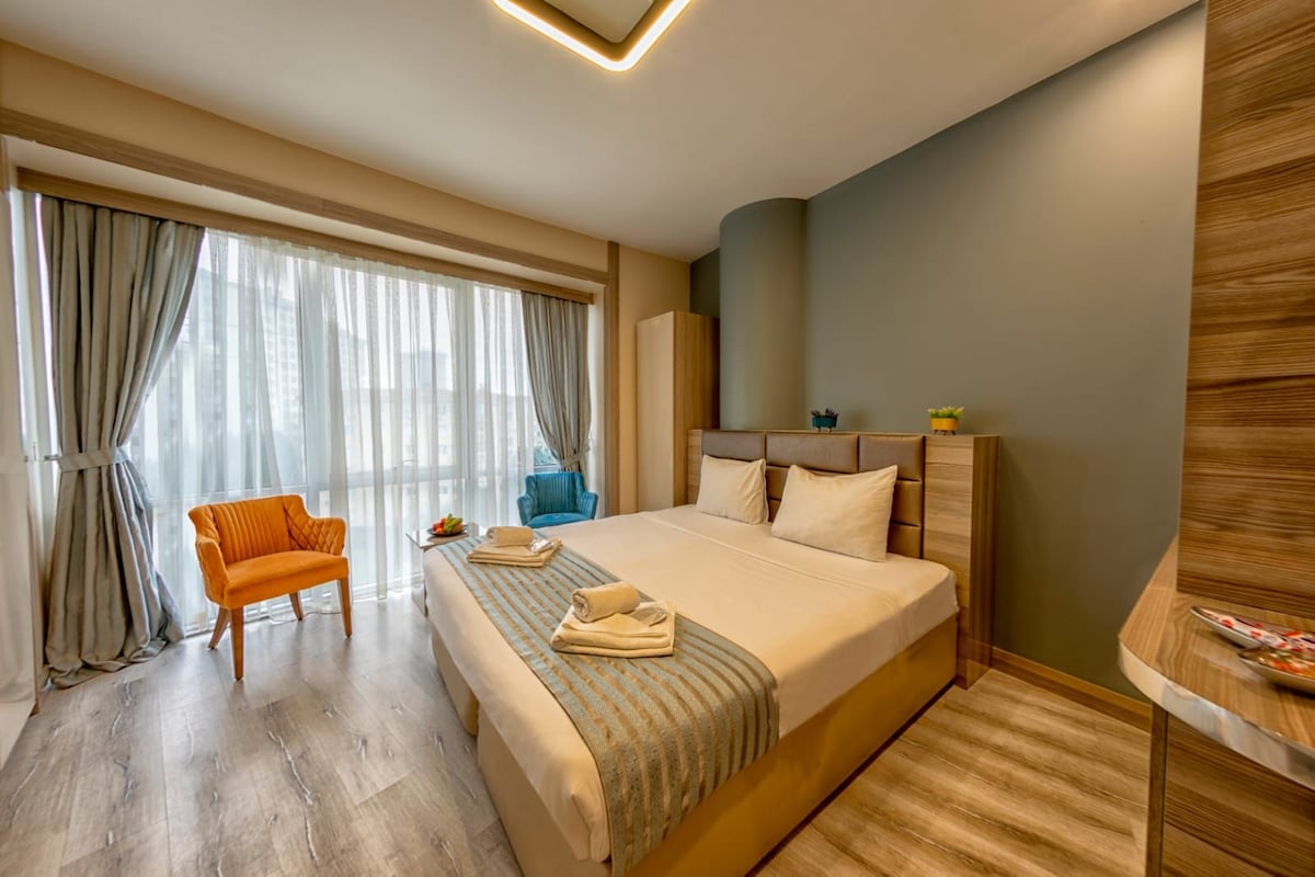 Quarto de hotel com cama grande, uma cadeira, mesa de cabeceira e janela com cortinas.