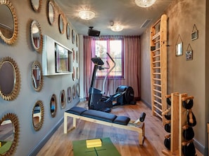 Quarto com esteira, banco de exercícios, espelhos fixados na parede, suporte de madeira e janela com cortinas.