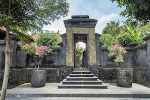 Entrada de um templo com um portal central, ladeado por grandes árvores e plantas em vasos.