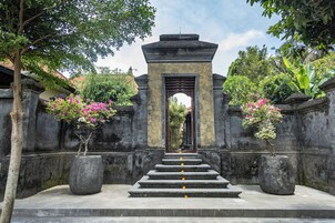 Entrada de um templo com um portal central, ladeado por grandes árvores e plantas em vasos.
