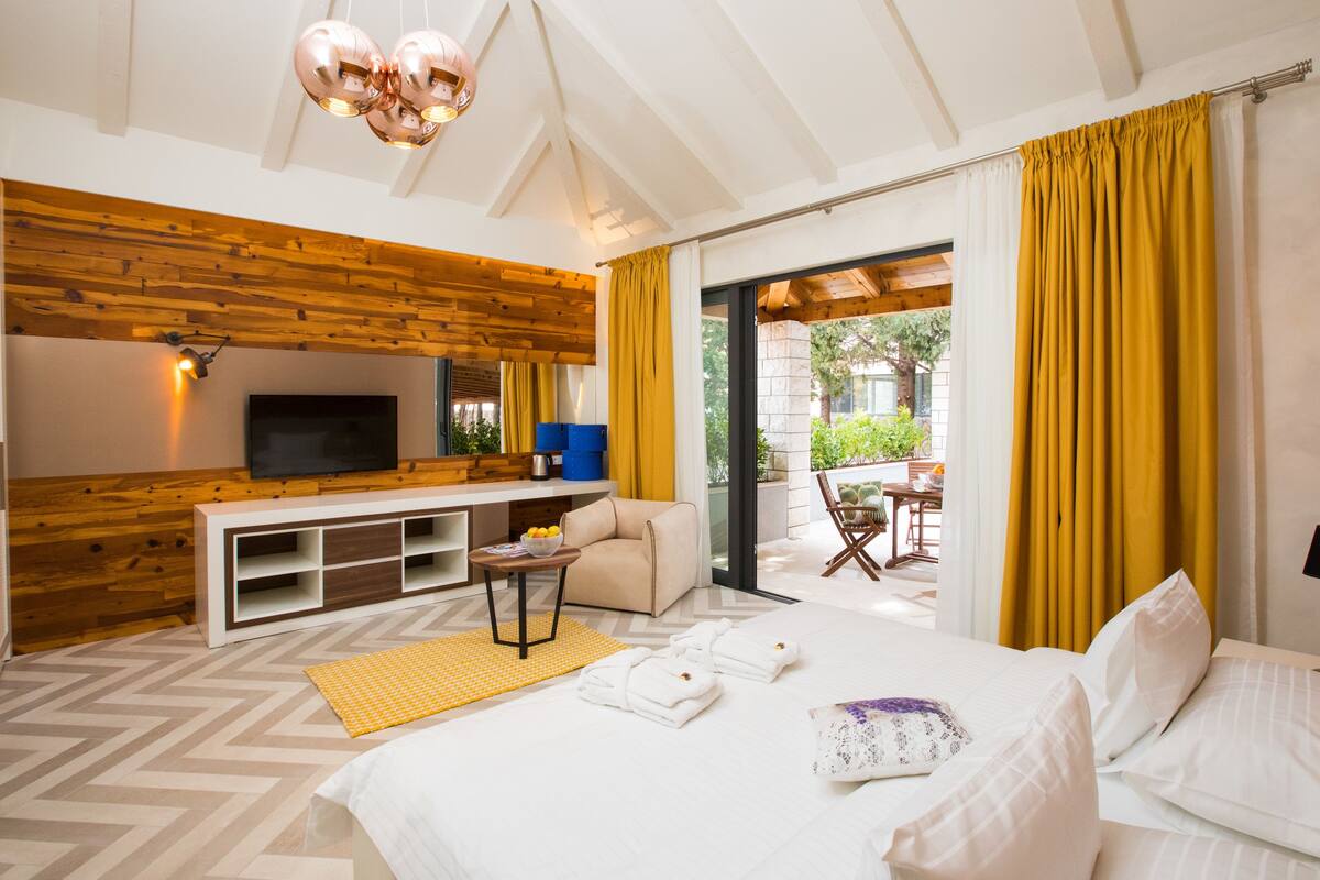 Quarto de hotel moderno com cama, televisão, parede de madeira, sofá e varanda com cortinas amarelas.