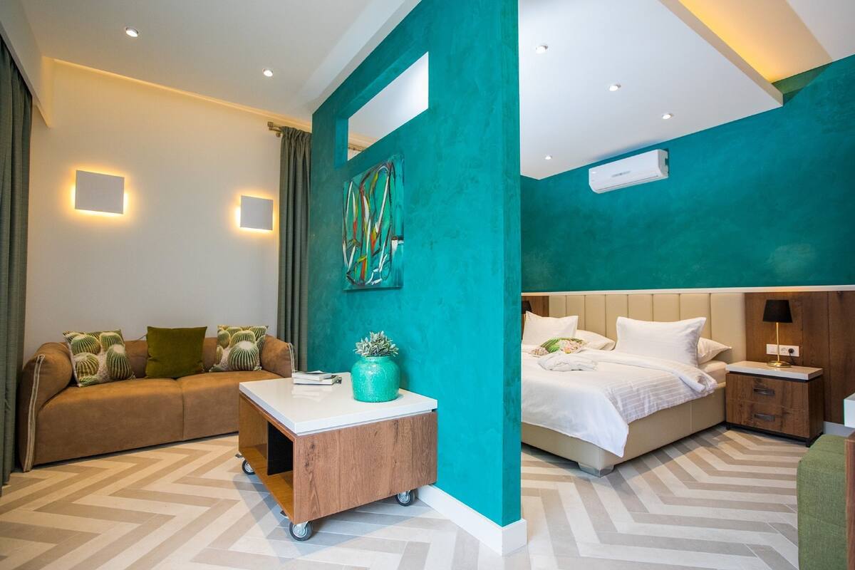 Quarto de hotel moderno com uma parede de destaque verde-azulado, sofá, cama, escrivaninha e uma planta pequena.