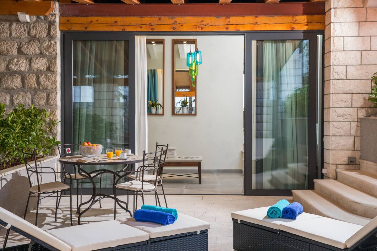 Um pátio externo com mesa de jantar, cadeiras e espreguiçadeiras. Há uma porta de correr de vidro que dá para uma área interna com uma mesa e cadeiras.
