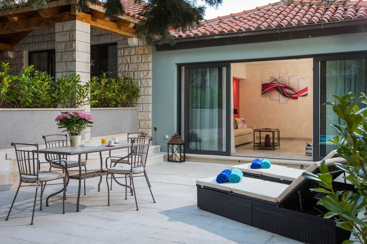 Um pátio externo com mesa e cadeiras de jantar, um espaço de descanso em vime e vista para o interior através de uma porta de vidro.
