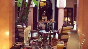 Mesa posta com pratos tradicionais marroquinos, um jarro de suco de laranja e um bule de chá, cercada por cadeiras e uma planta decorativa.