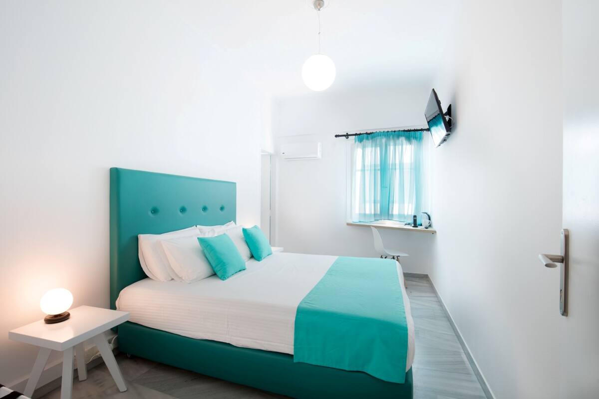 Quarto com cama verde-água, roupa de cama branca, mesa de cabeceira com abajur, janela com cortinas e televisão presa na parede.