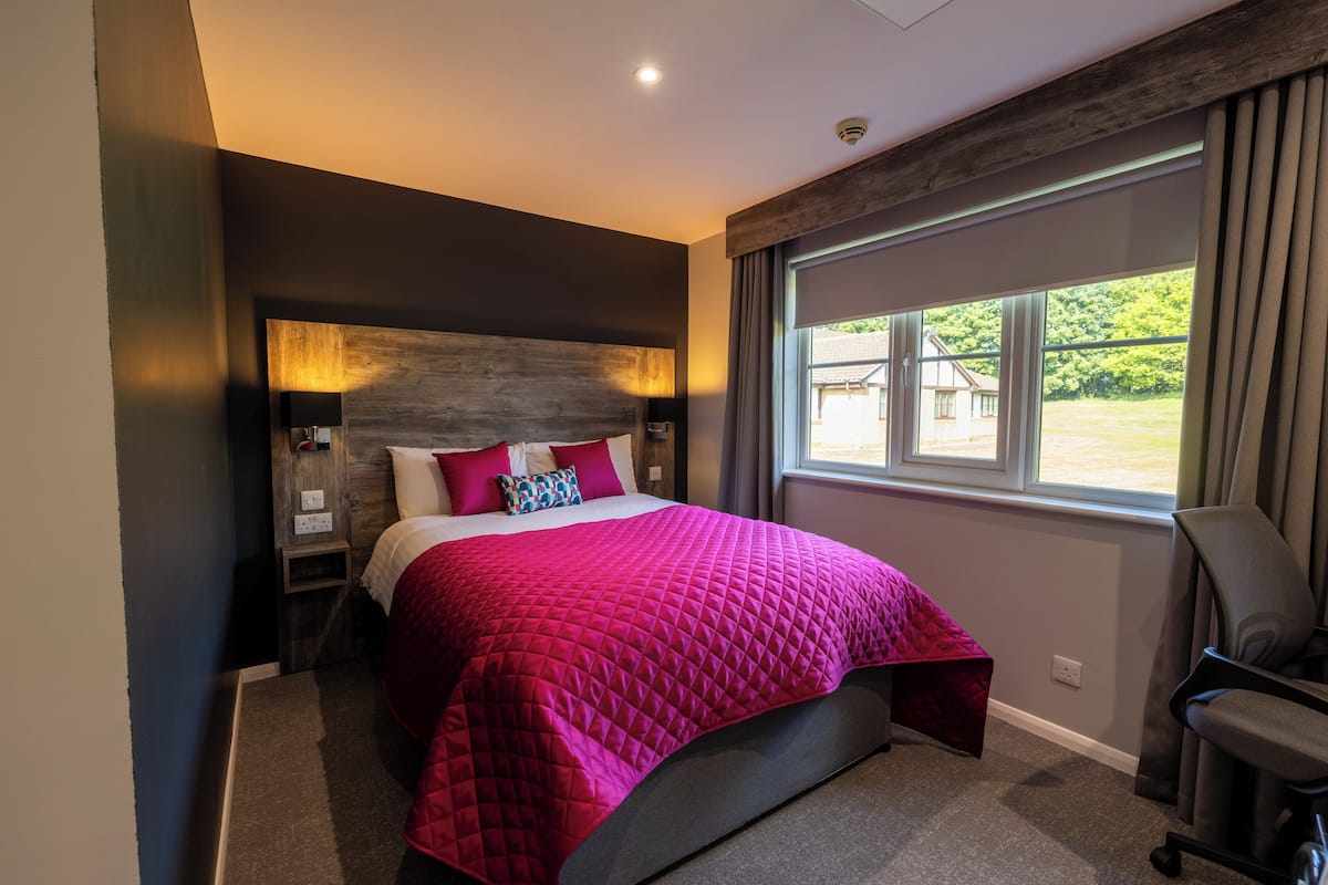 Quarto moderno com uma cama grande, cabeceira, janela com cortinas e uma escrivaninha com cadeira.