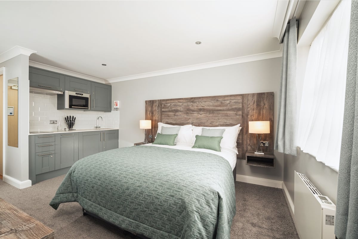 Quarto com cama grande, cabeceira de madeira, duas luminárias de apoio e uma kitchenette com armários cinzentos e micro-ondas.
