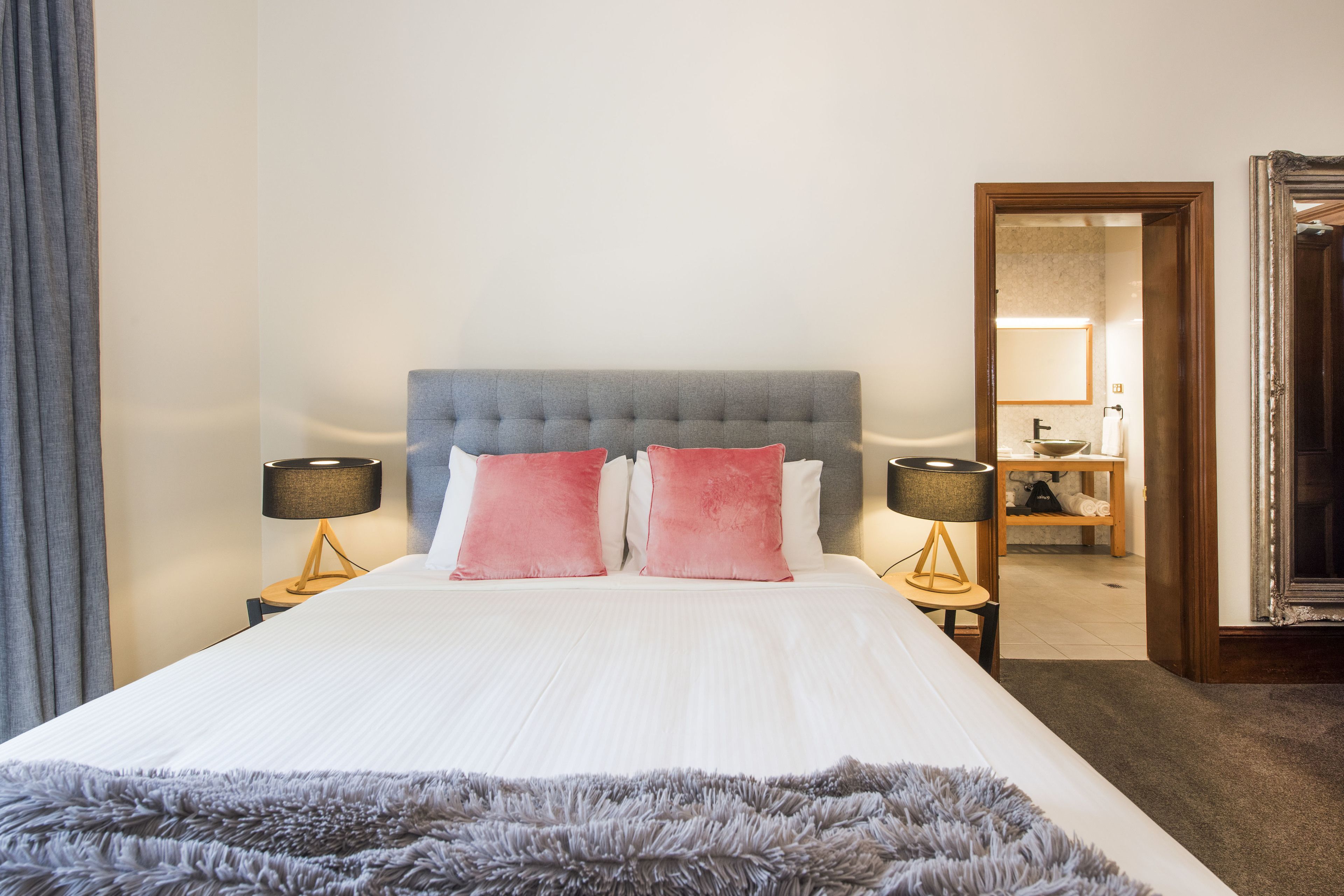 Cama bem arrumada com cabeceira cinza, duas almofadas cor-de-rosa e um cobertor felpudo cinza. Duas luminárias de cabeceira com cúpulas em cada lado da cama.