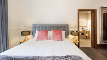 Cama bem arrumada com cabeceira cinza, duas almofadas cor-de-rosa e um cobertor felpudo cinza. Duas luminárias de cabeceira com cúpulas em cada lado da cama.