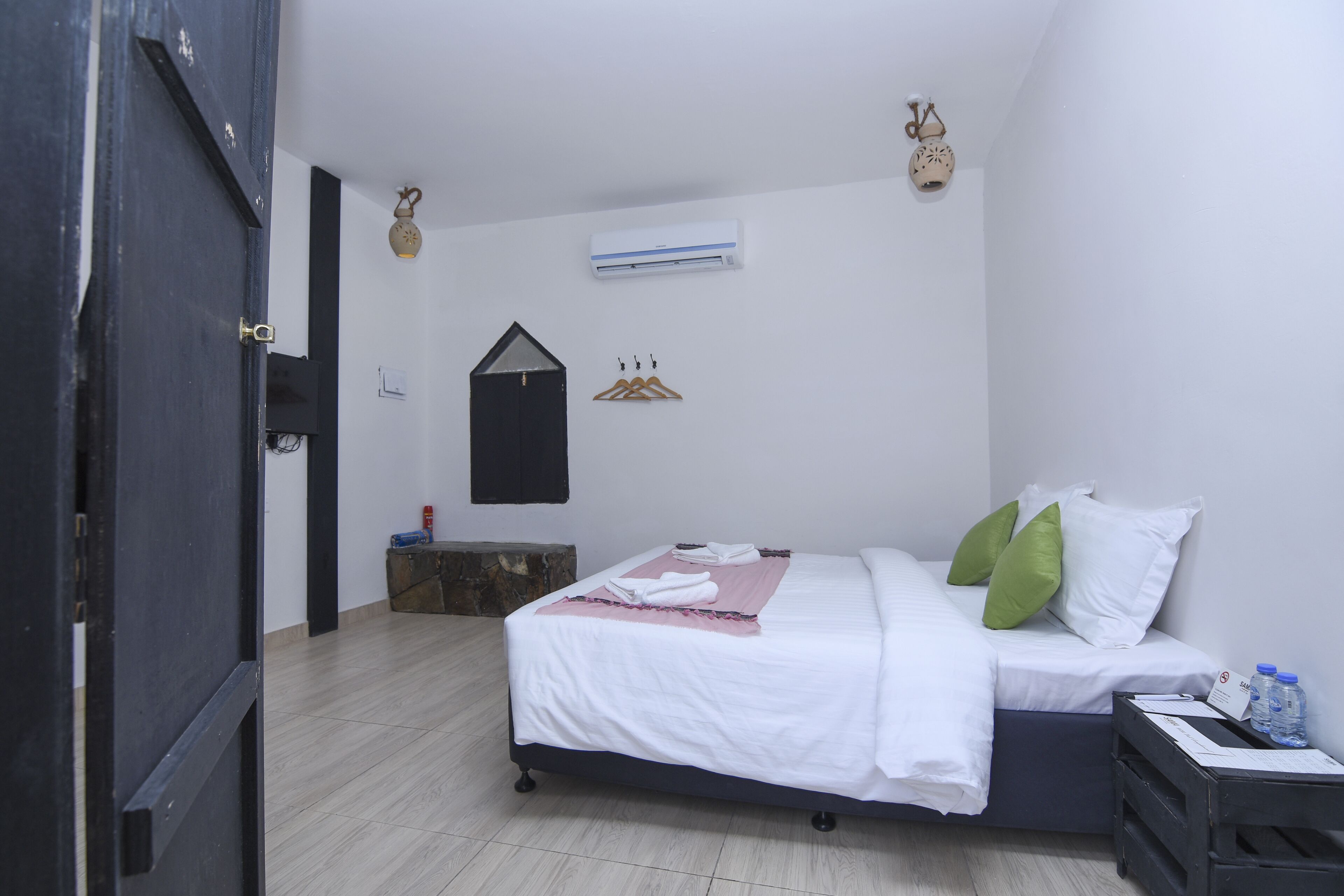 Quarto de hotel com uma cama, uma mesa pequena, uma mesinha de cabeceira, ar condicionado, um cabideiro fixado na parede e uma porta.