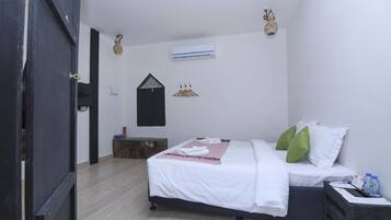 Quarto de hotel com uma cama, uma mesa pequena, uma mesinha de cabeceira, ar condicionado, um cabideiro fixado na parede e uma porta.