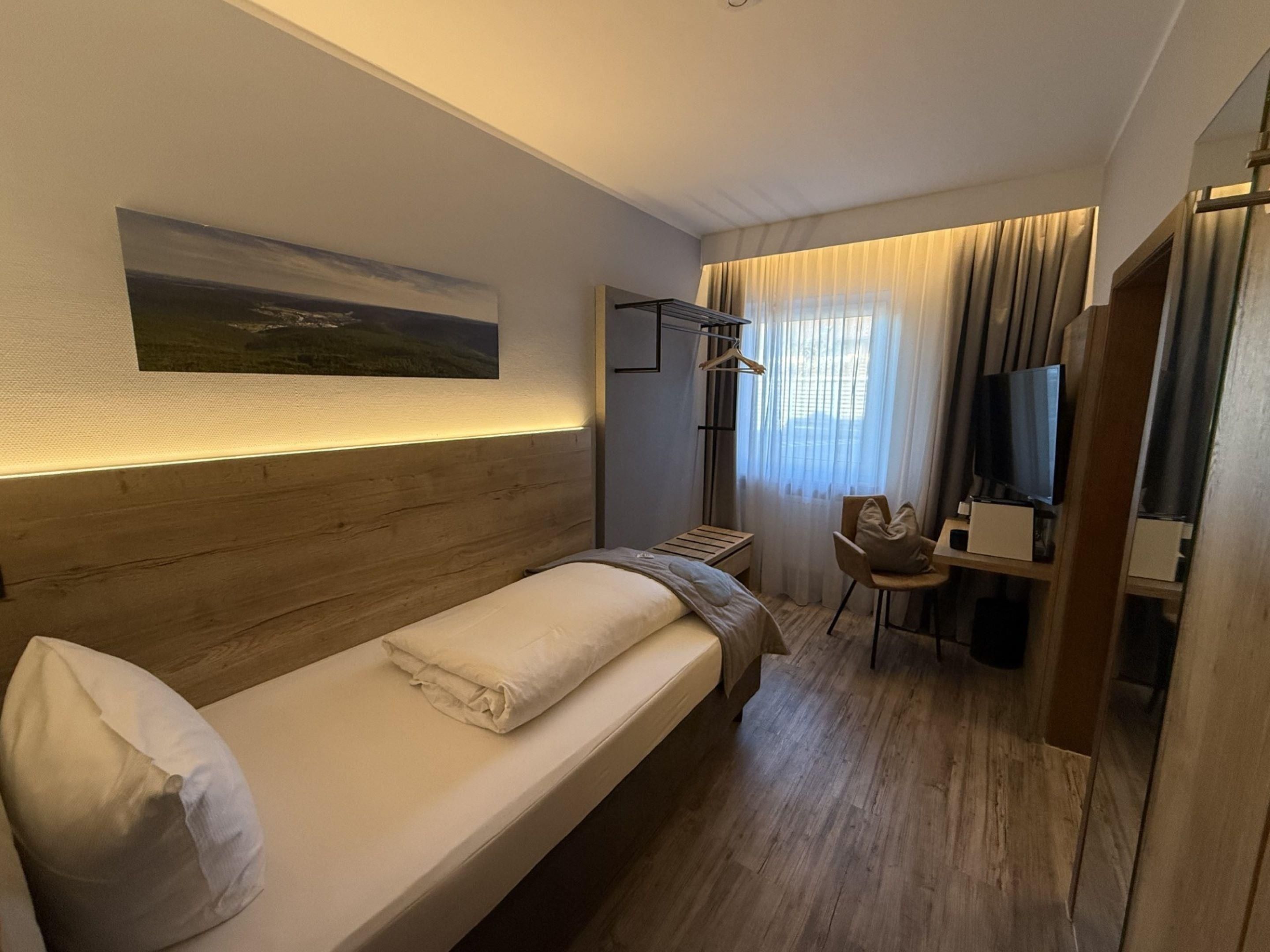 Quarto de hotel com cama, escrivaninha, cadeira, televisão e um quadro na parede.