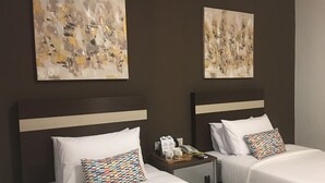 Duas camas de casal com roupa de cama branca, uma pequena mesa de cabeceira com um telefone e uma chávena, e quadros abstratos nas paredes.