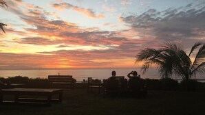 Vista do pôr do sol a partir de uma área de estar à beira-mar, com bancos e palmeiras.