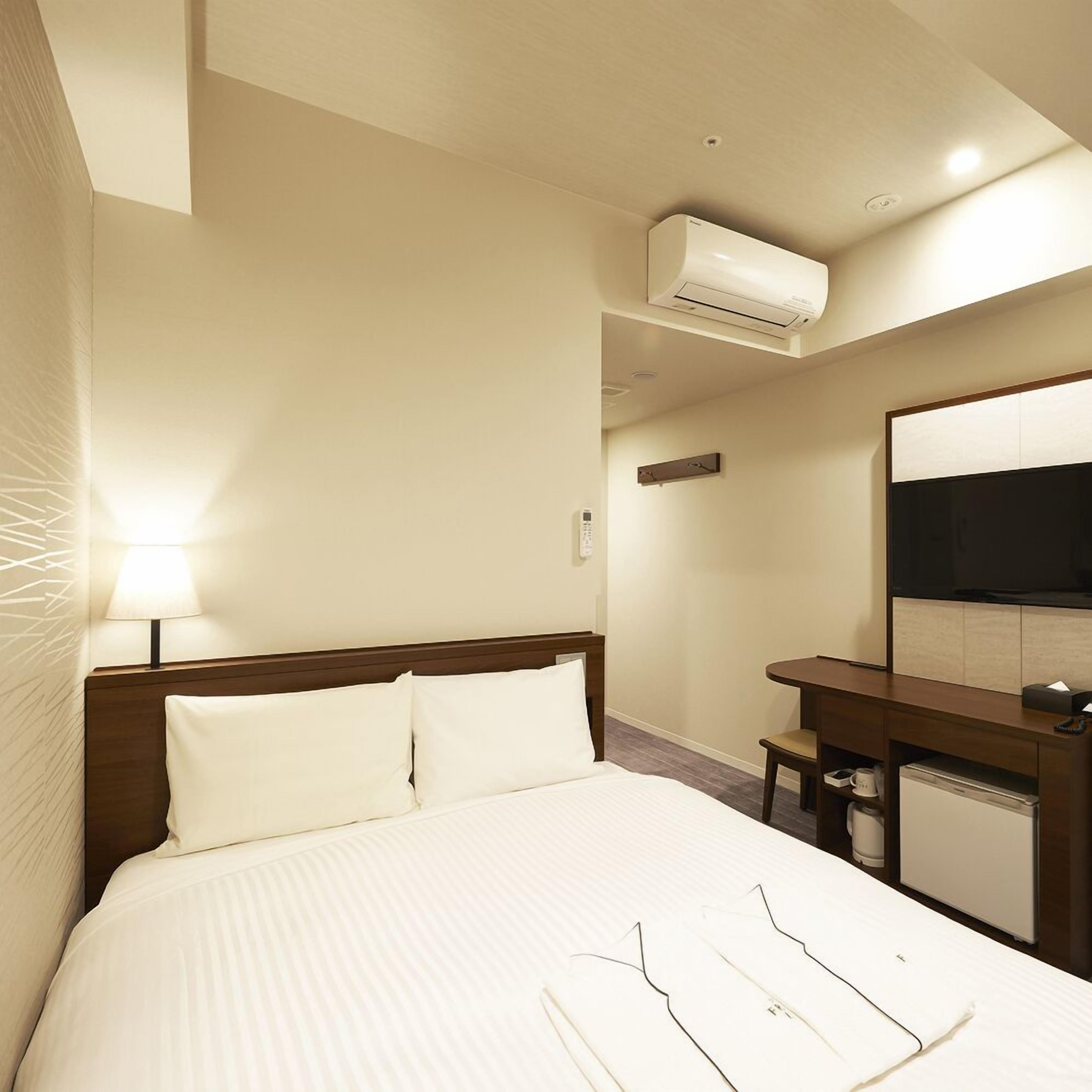 Quarto de hotel com cama, luminária, ar condicionado, escrivaninha, cadeira e televisão.