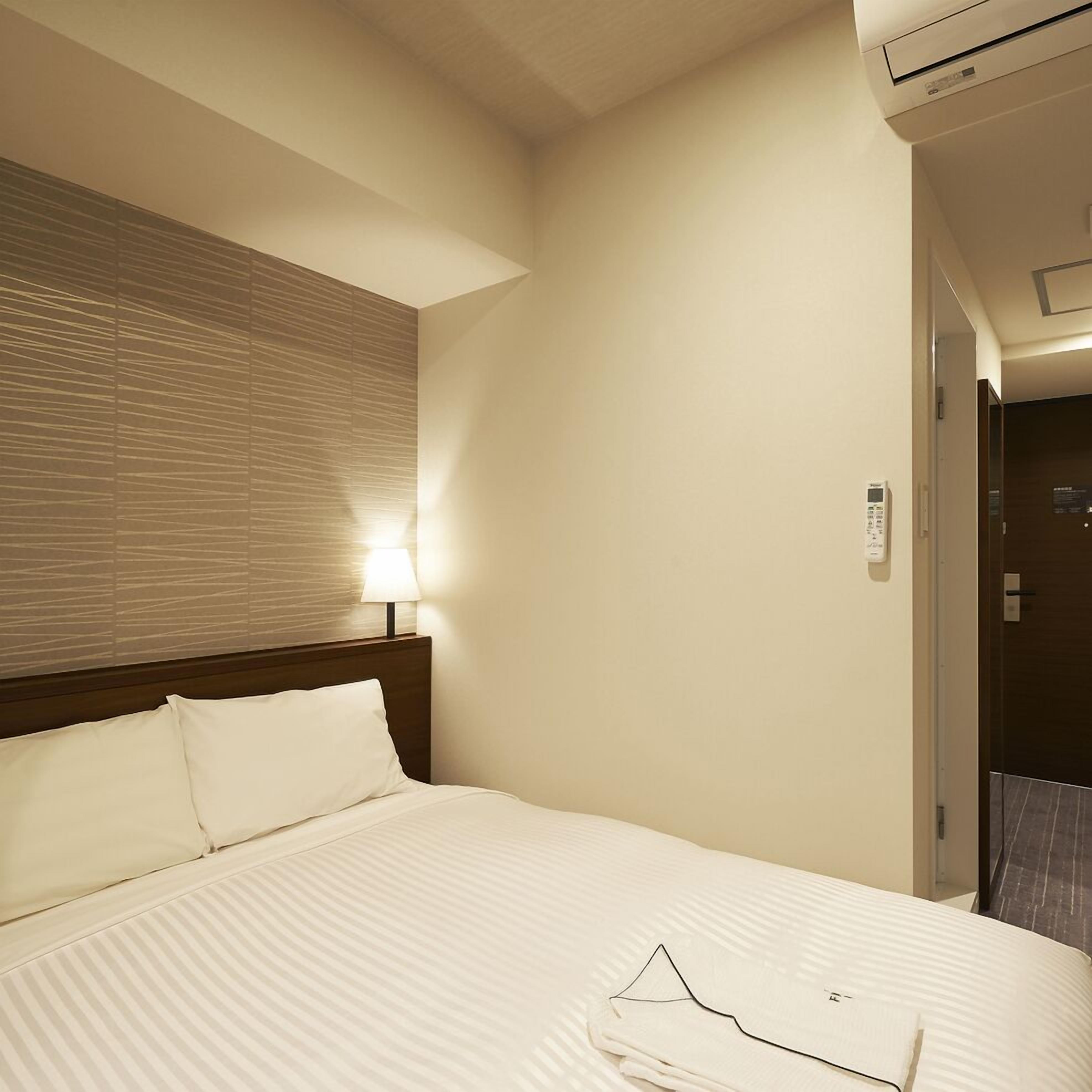 Quarto de hotel com uma cama, uma mesinha de cabeceira com abajur, ar-condicionado fixado na parede e uma porta que dá para outro cômodo.