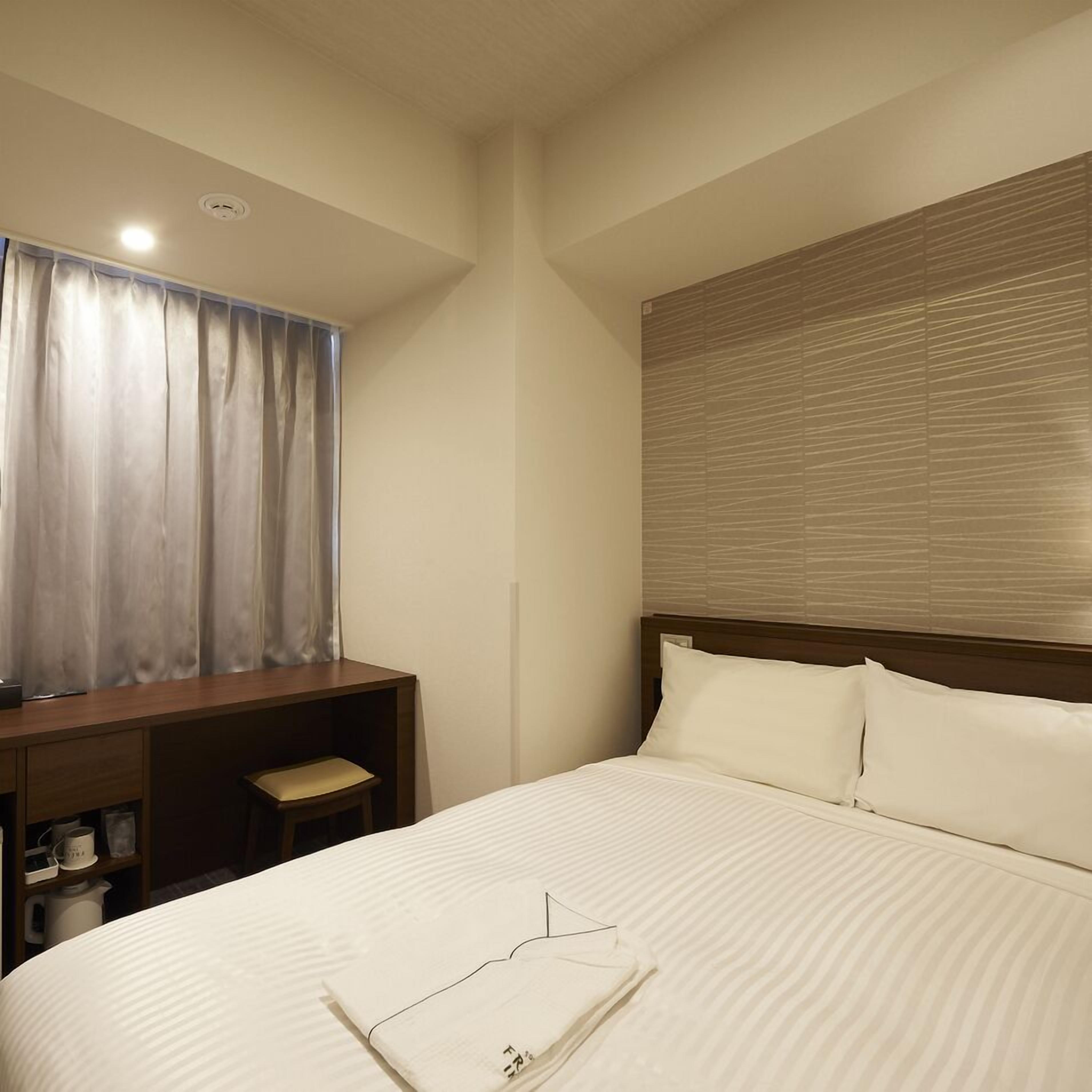 Quarto de hotel com cama, escrivaninha, cadeira e janela com cortinas.