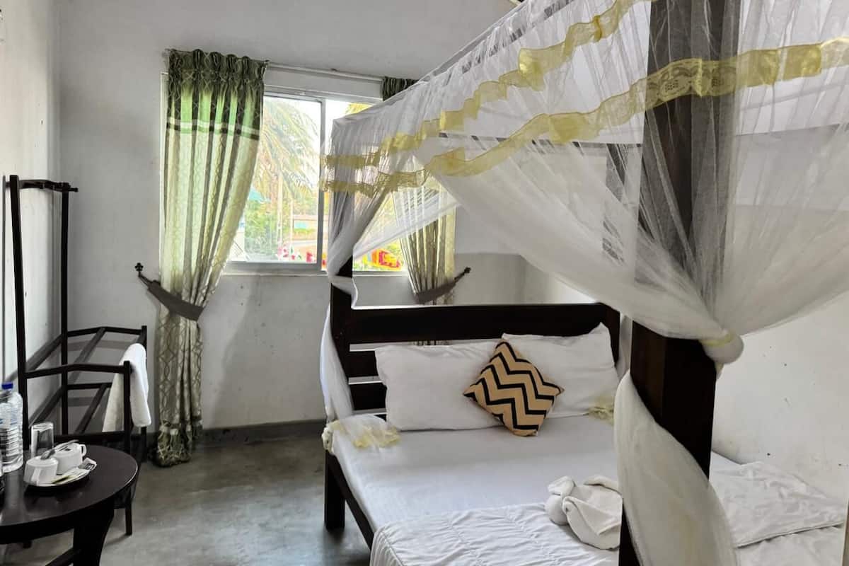 Quarto com cama, uma mesinha com cadeira, janela com cortinas e vista para o exterior.