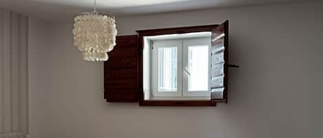 Quarto com cama, cadeira, mesa pequena, janela com cortinas e um candelabro.