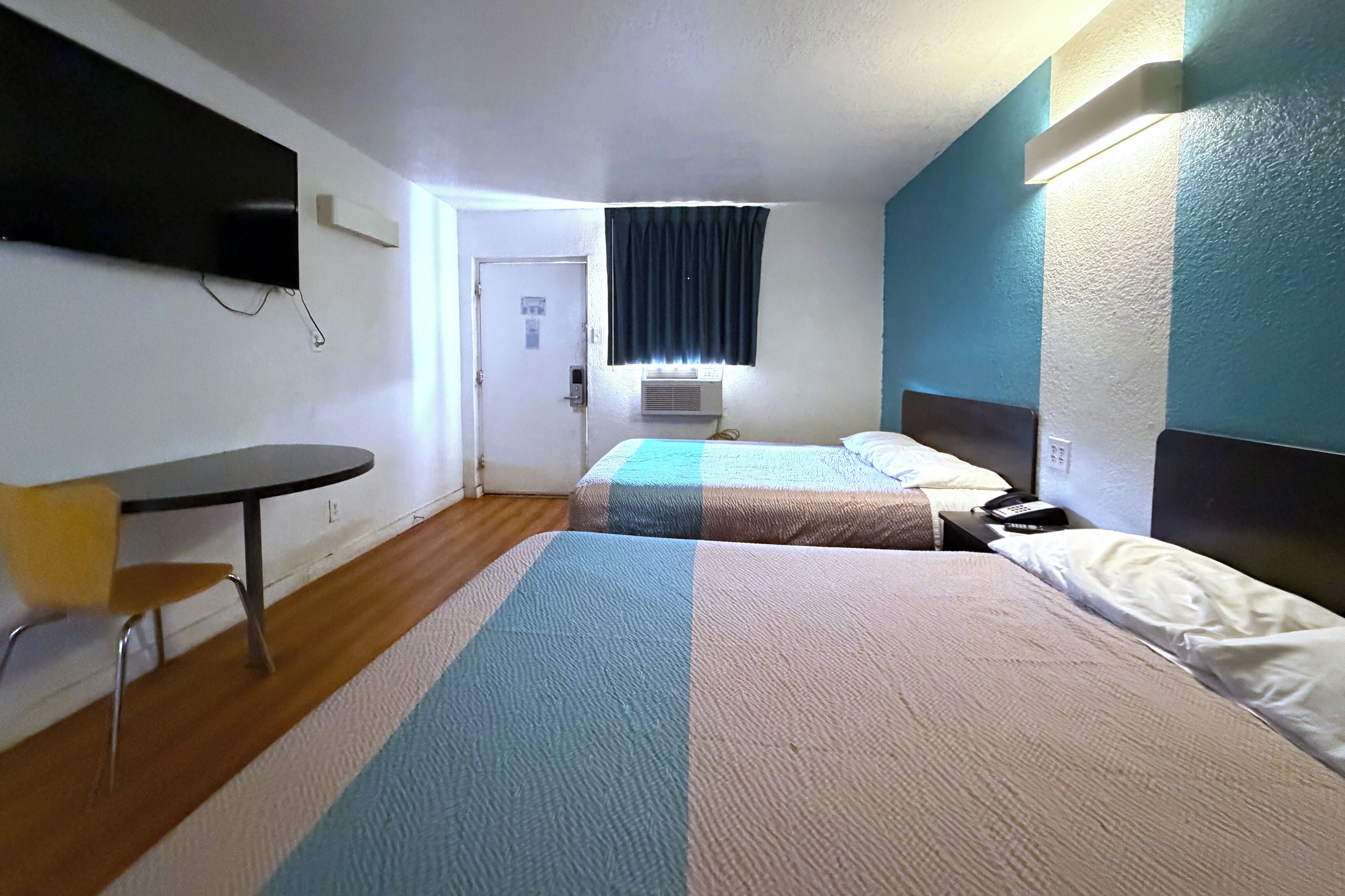 Quarto de hotel com cama, uma cadeira, uma mesa, TV fixada na parede e janela com cortinas.