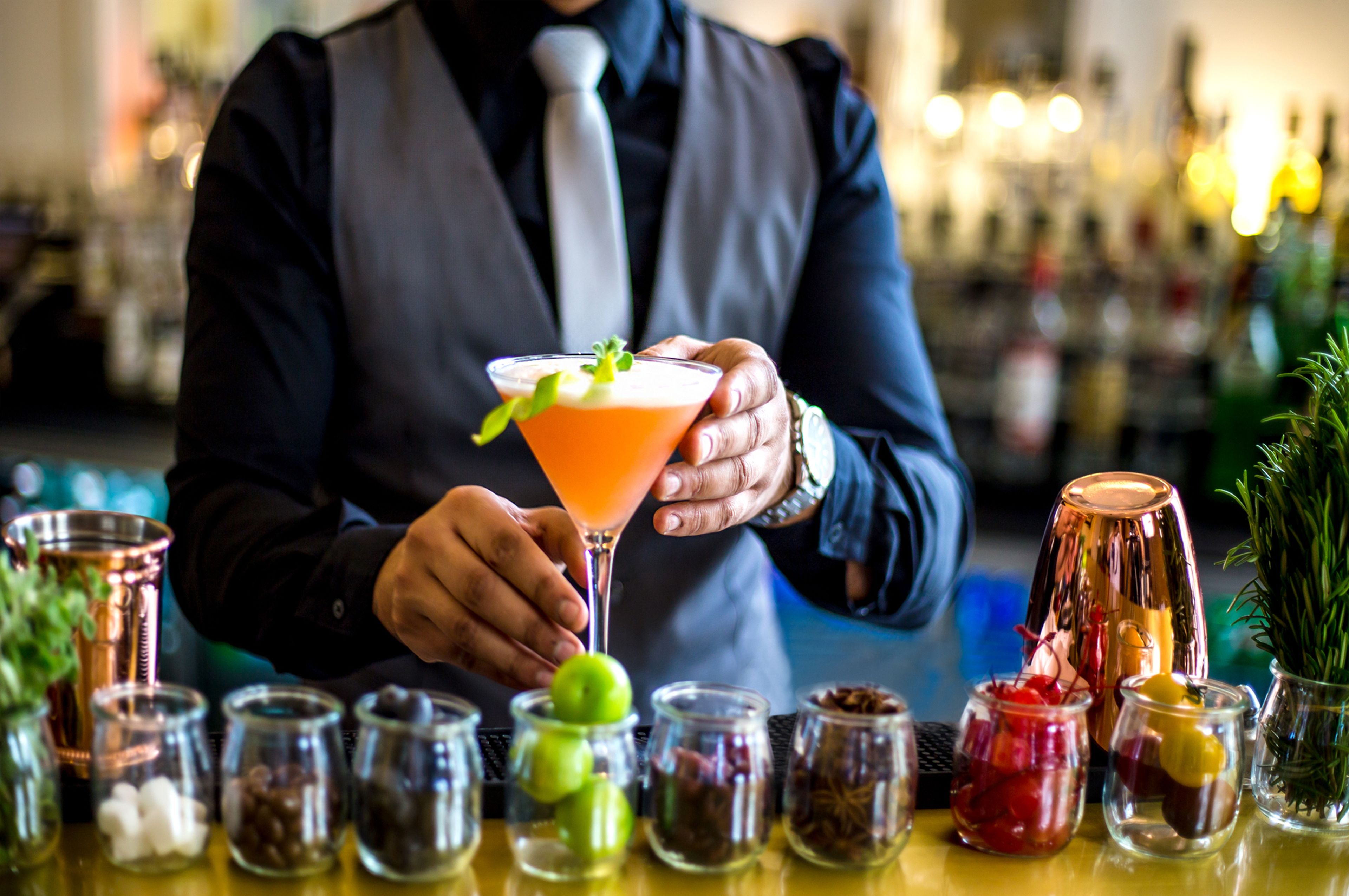 Um bartender segurando um coquetel com diversos enfeites e ingredientes em um balcão.