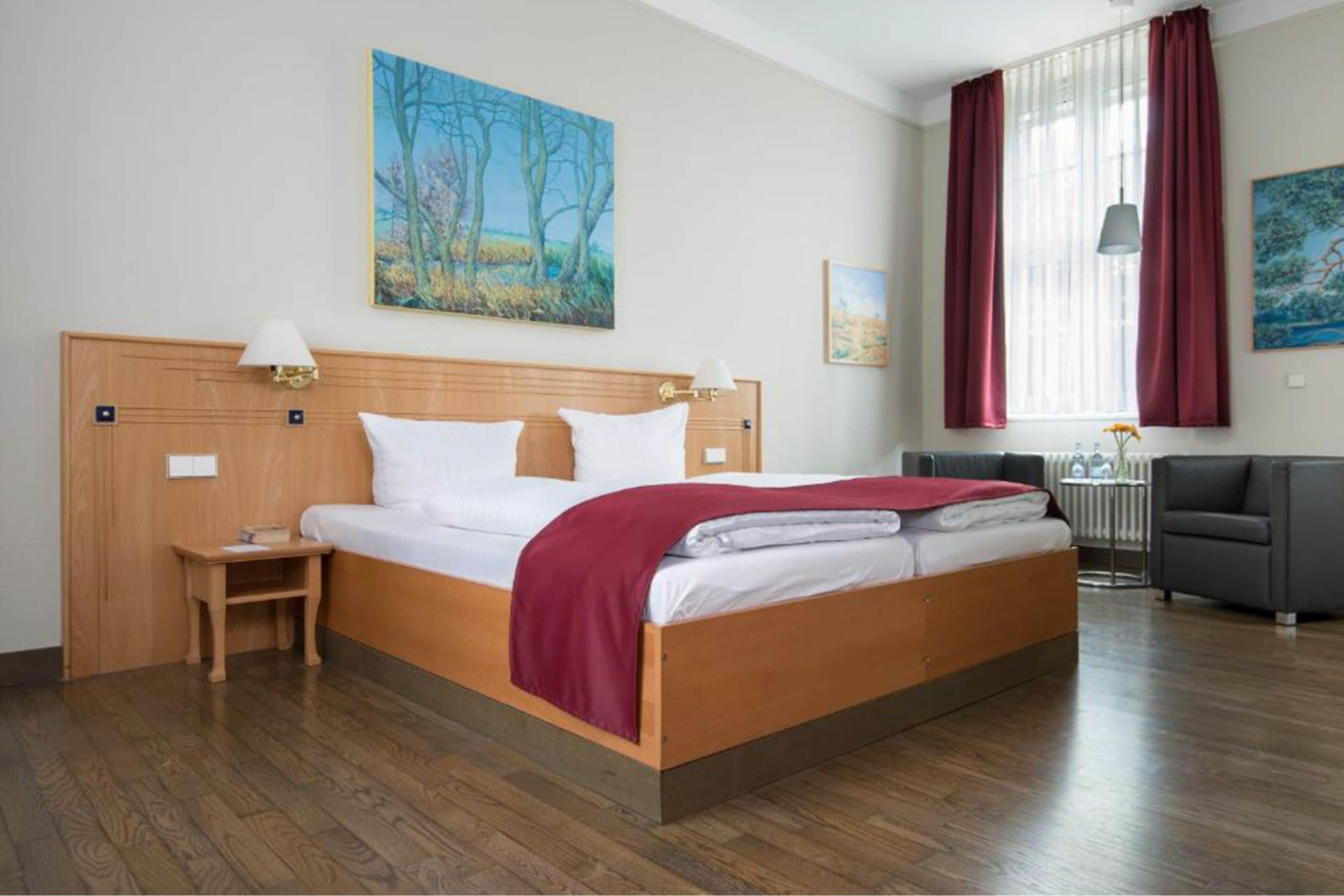 Quarto de hotel com cama de madeira, mesa de cabeceira, sofá e duas pinturas nas paredes.