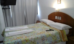Quarto de hotel com cama, televisão presa à parede e janela com cortinas.