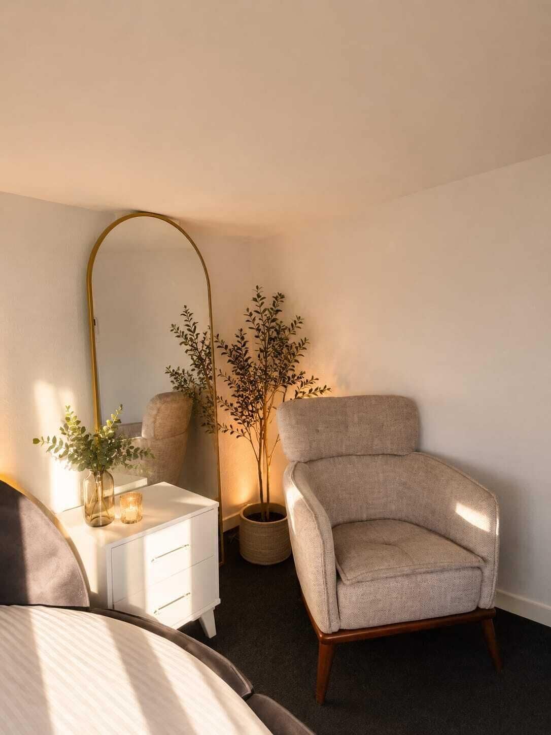 Quarto com cama, uma cadeira, mesa de cabeceira com luminária, espelho e planta em vaso.